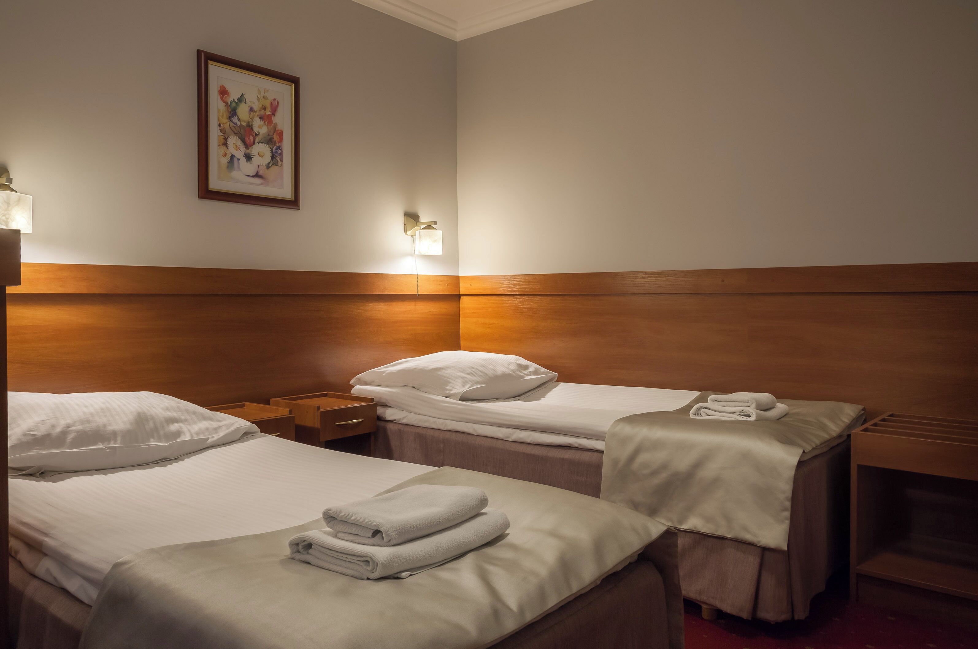 Quarto de hotel com duas camas, uma cabeceira de madeira, uma mesinha de cabeceira, uma obra de arte floral emoldurada e uma luminária fixada na parede.