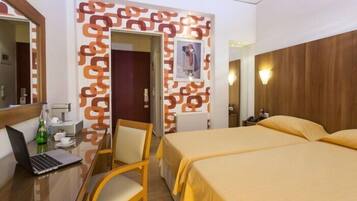 Quarto de hotel com uma cama, uma escrivaninha com um laptop, uma cadeira, um espelho e uma parede com padrão geométrico.