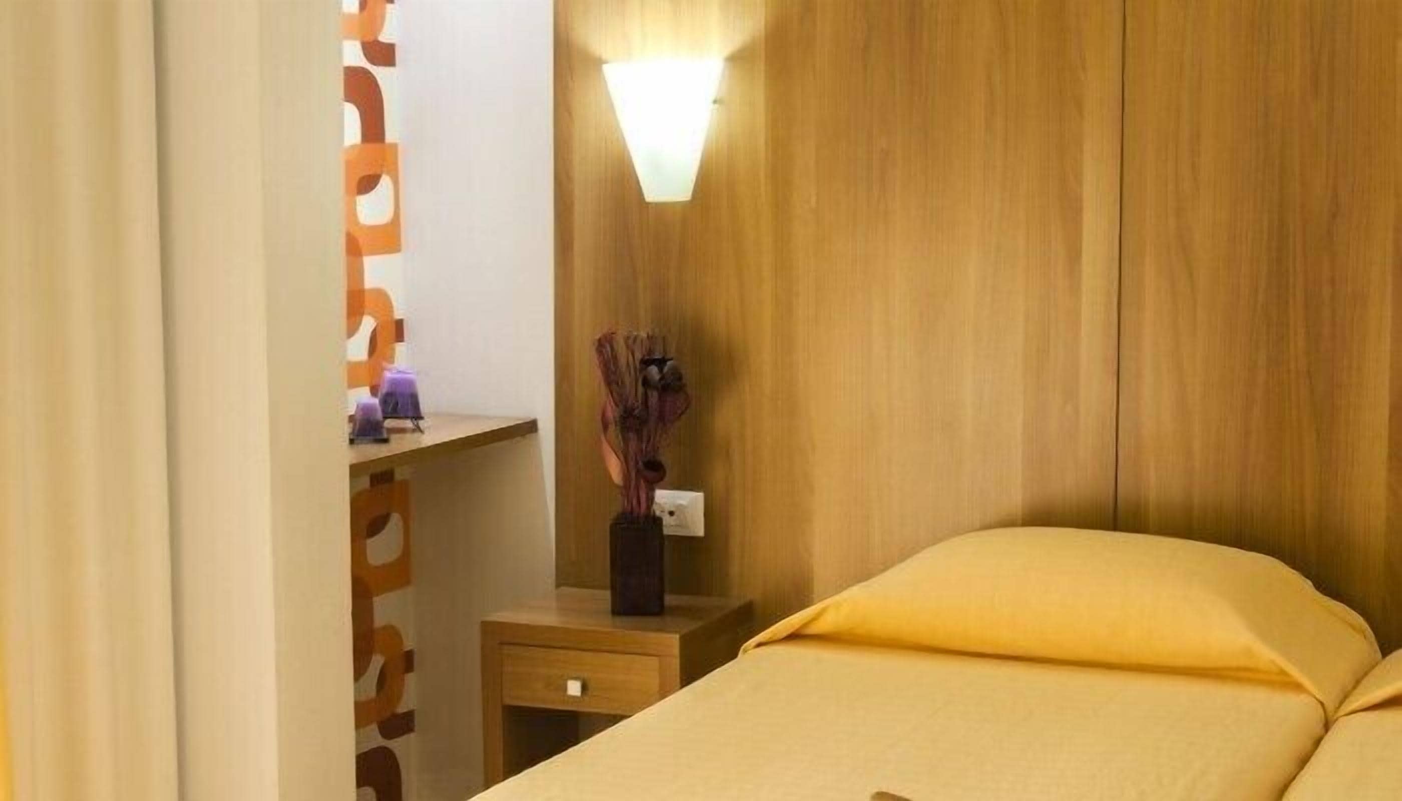 Quarto de hotel com guarda-roupa de madeira, mesa de cabeceira com um vaso de flores secas e uma cama com colcha amarela.