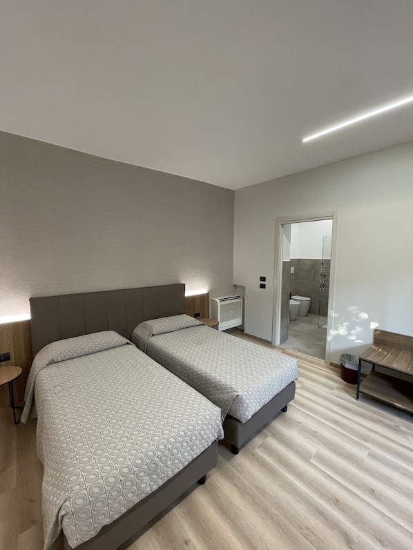 Quarto de hotel moderno com duas camas, uma mesinha, um banco e um banheiro visível através de uma porta de vidro.