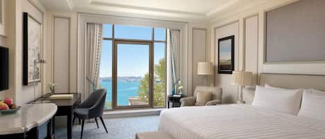 Quarto de hotel com uma cama grande, uma escrivaninha, uma cadeira, vista para o mar e para a cidade.