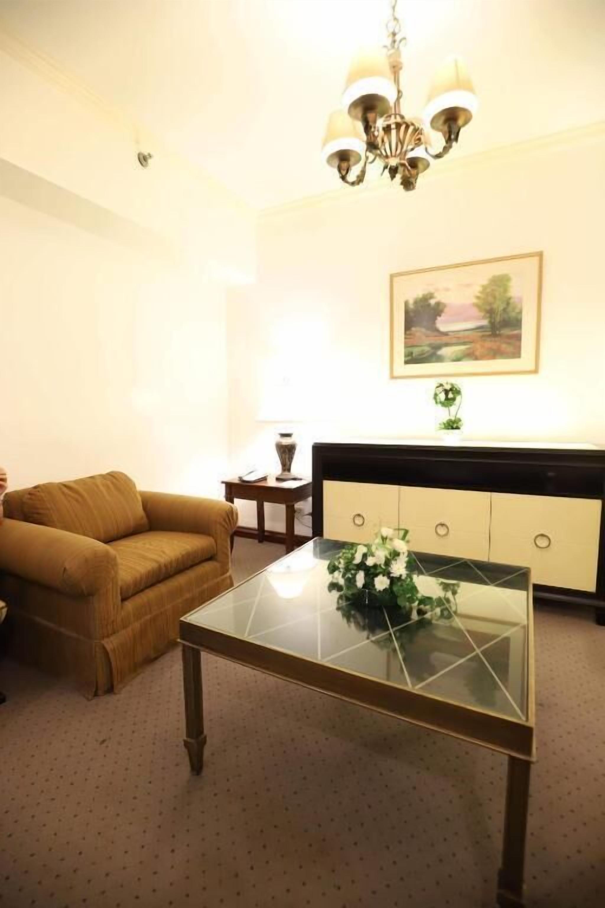 Uma sala de estar com sofá, mesa de centro de vidro, console com gavetas, abajur e um quadro na parede.