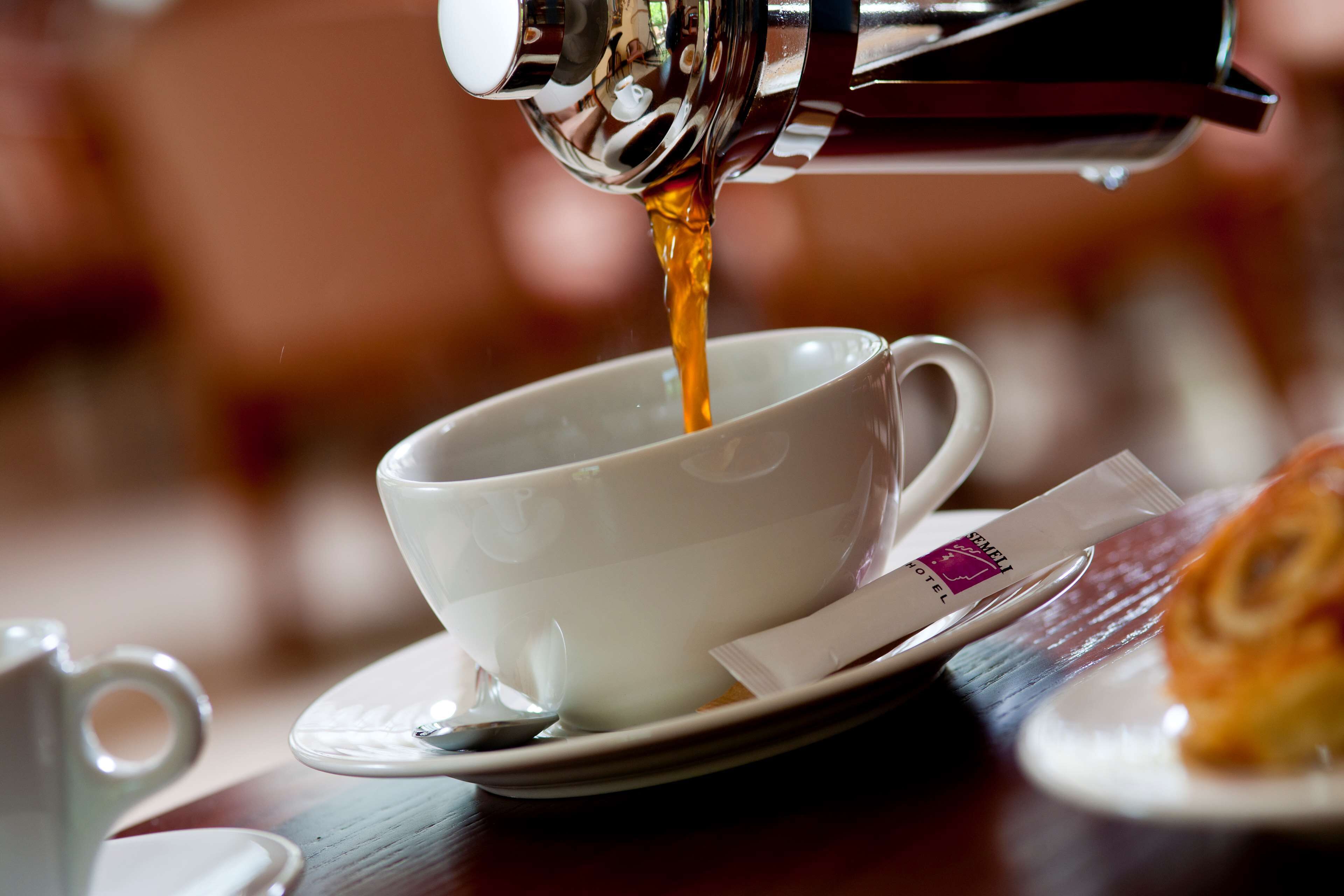 Uma chávena de café a ser servida numa chávena branca sobre um pires, com um pastel e um saquinho de chá na mesa.