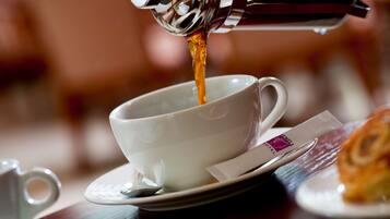 Uma chávena de café a ser servida numa chávena branca sobre um pires, com um pastel e um saquinho de chá na mesa.