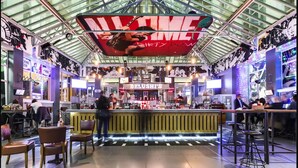Um bar em ambiente interno, com teto alto, iluminação neon e balcão central.