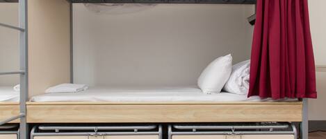 Uma cama beliche com estrutura de metal, roupa de cama branca e cortina vermelha. Há gavetas para guardar objetos embaixo da cama.