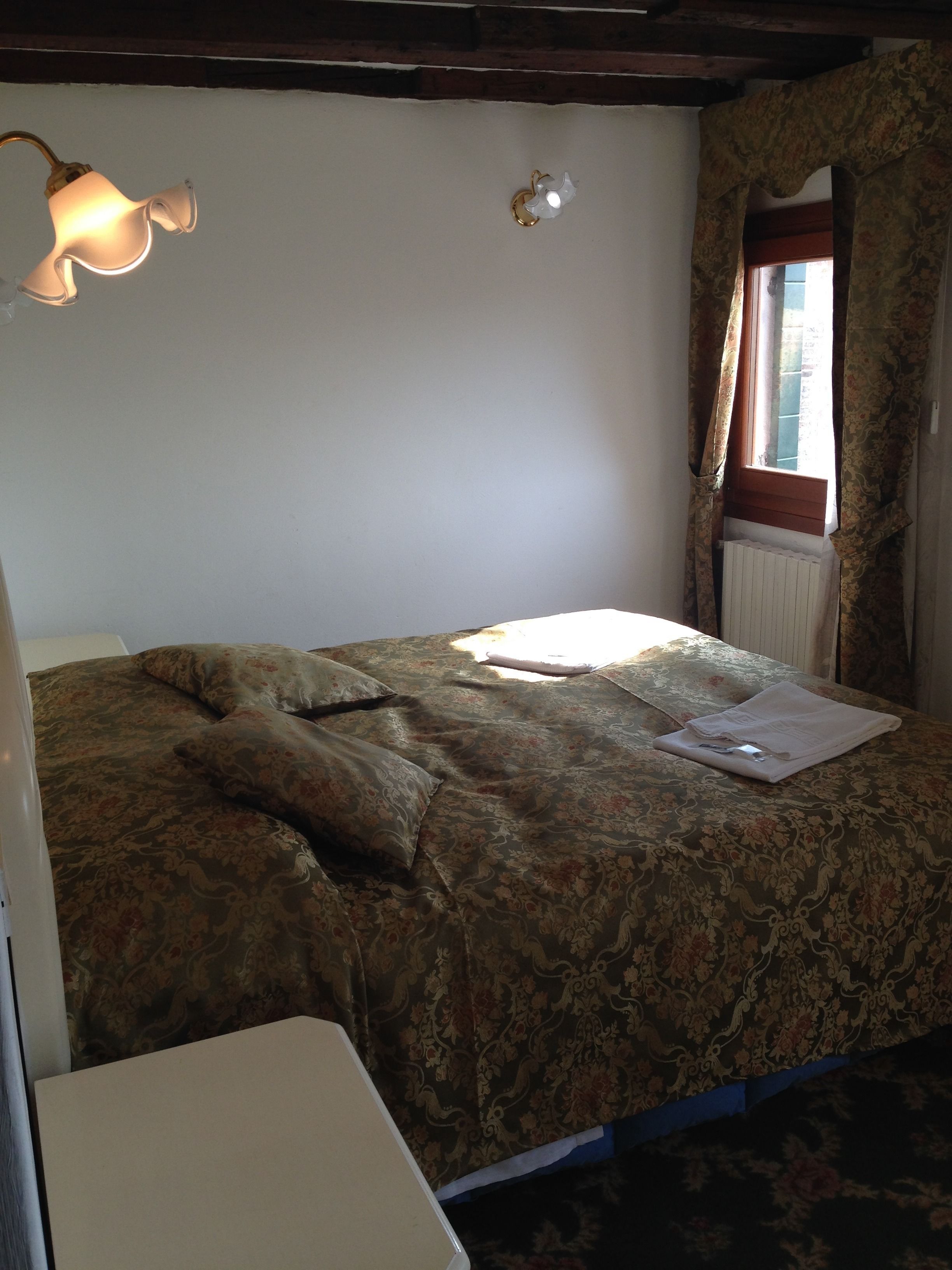 Um quarto com uma cama, uma mesa pequena, uma janela com cortinas e uma luminária na parede.