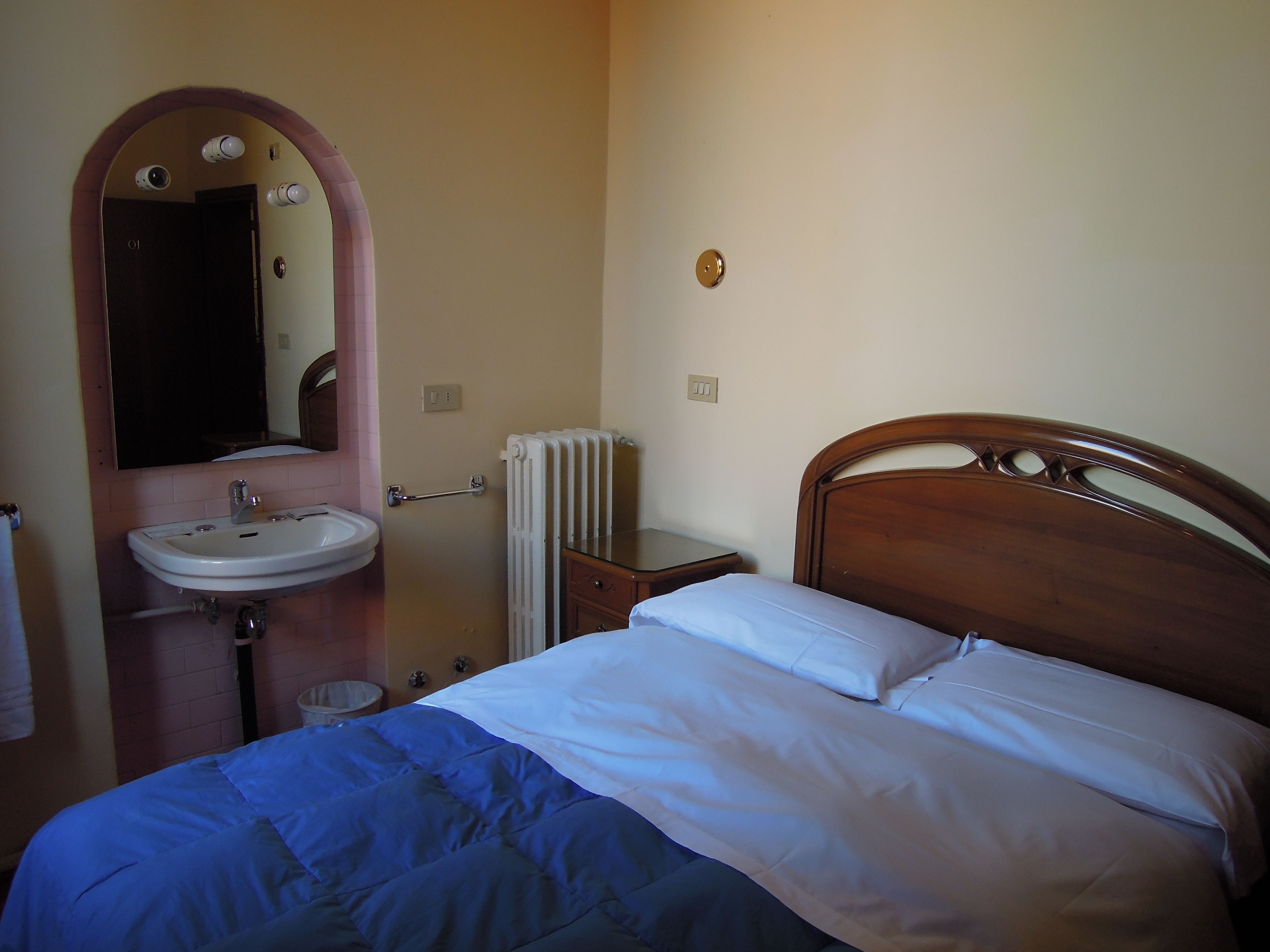 Quarto com cama, cabeceira de madeira, mesa de cabeceira, abajur branco, espelho, pia e radiador.