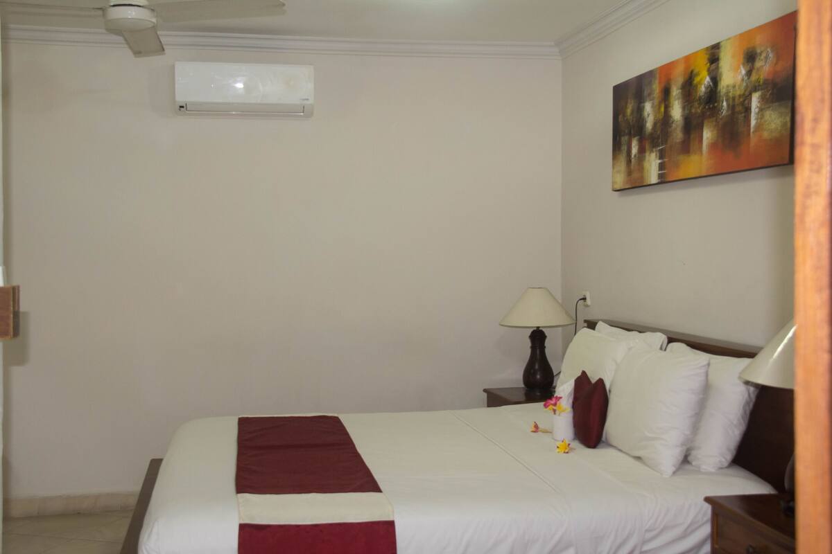 Quarto de hotel com cama, mesa de cabeceira com abajur, um quadro na parede e um ar condicionado.