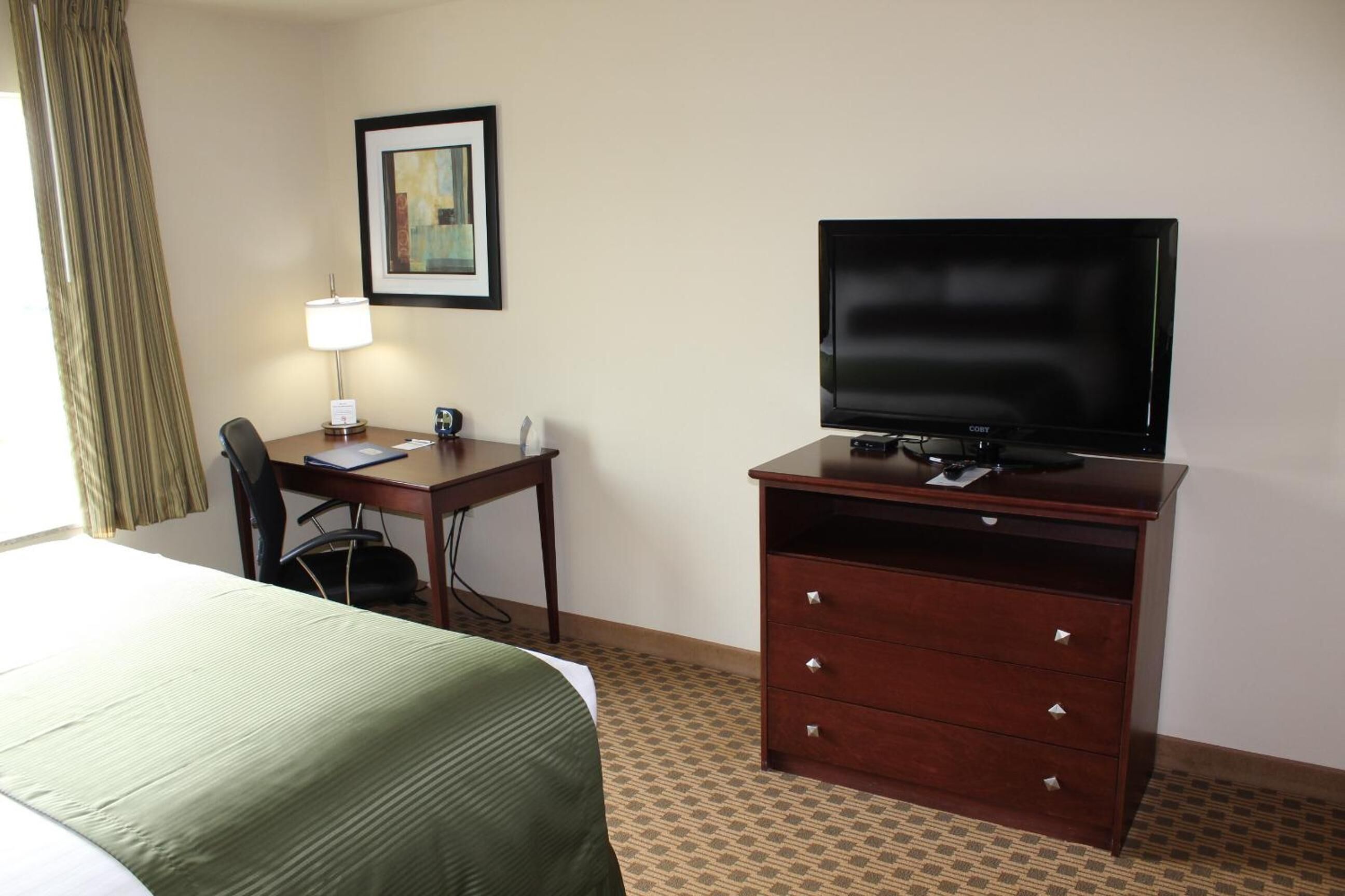 Quarto de hotel com cama, escrivaninha com abajur, televisão e cómoda.