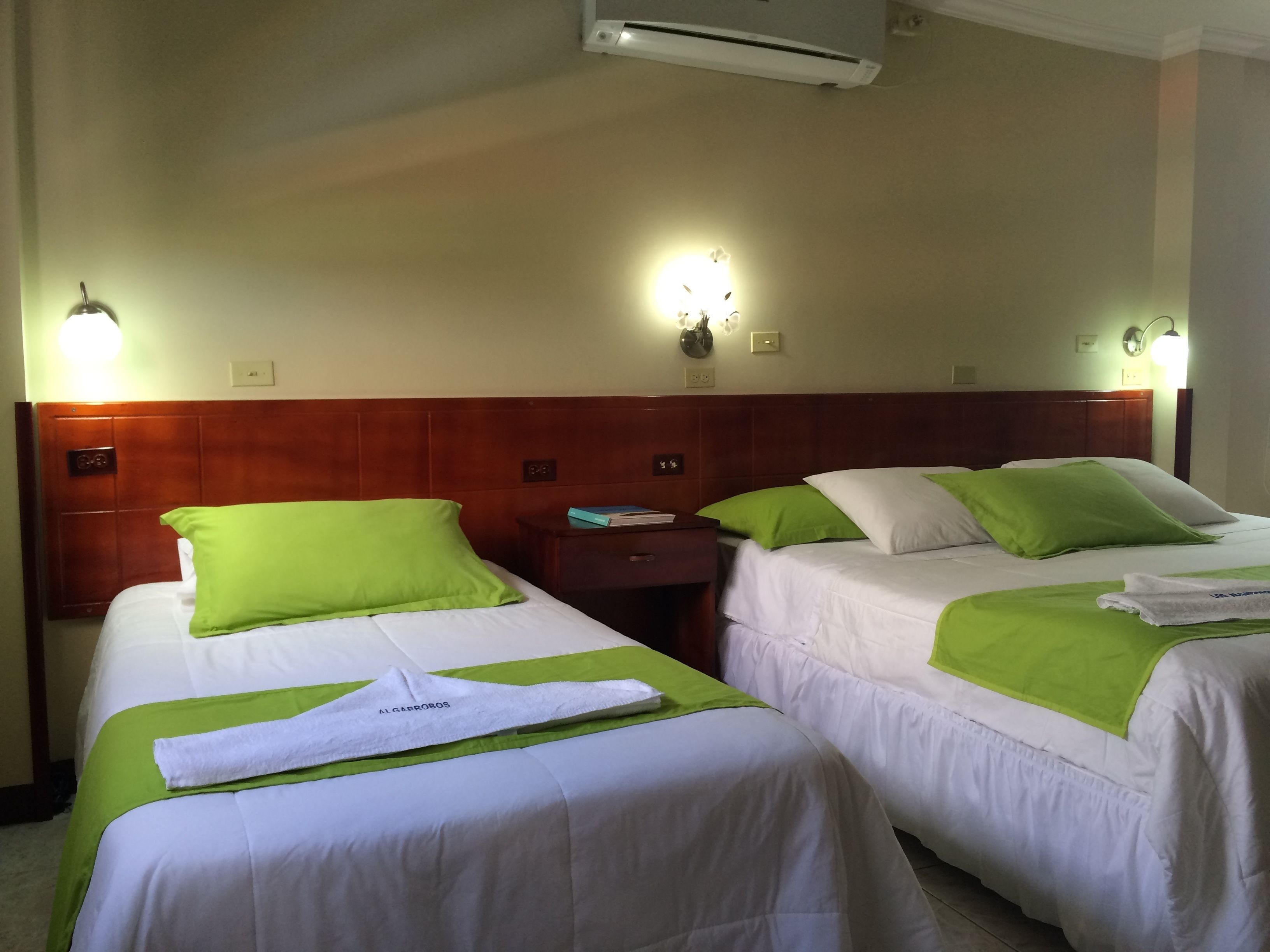 Duas camas de solteiro com roupa de cama verde e branca, cabeceira de madeira, mesas de cabeceira com luminárias e um aparelho de ar condicionado acima.