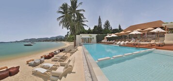 Uma área na beira da piscina com espreguiçadeiras e guarda-sóis, uma praia com palmeiras e vista para o oceano e montanhas ao longe.