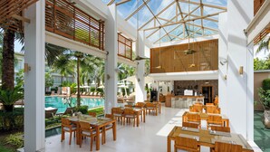 Uma ampla área de refeições interna com mesas e cadeiras de madeira, piscina visível através de grandes janelas e uma cozinha ao fundo.