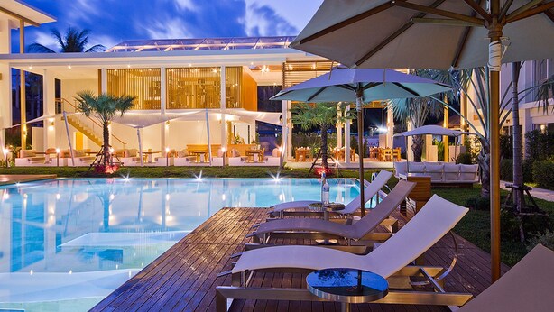 Área da piscina com espreguiçadeiras e guarda-sóis, um deck de madeira e um edifício moderno ao fundo.