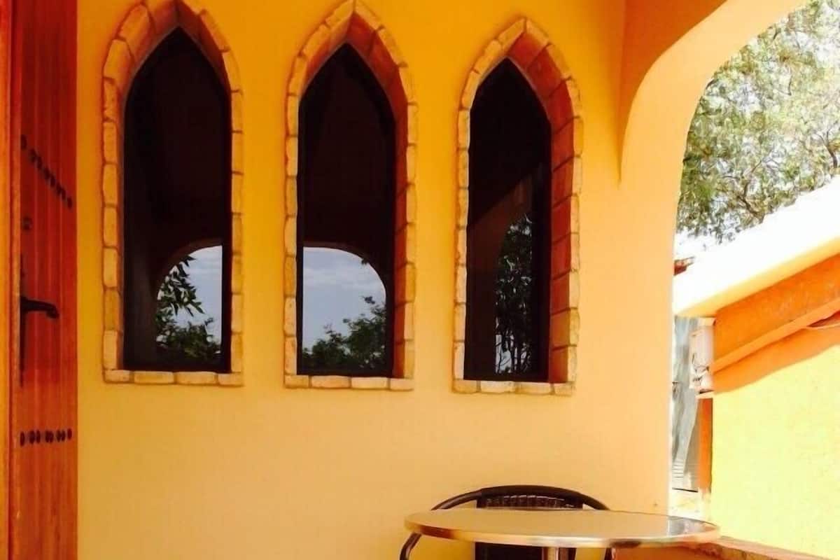 Um pequeno pátio com uma mesa redonda e duas cadeiras, piso de azulejos e três janelas arqueadas com um candeeiro em cima.