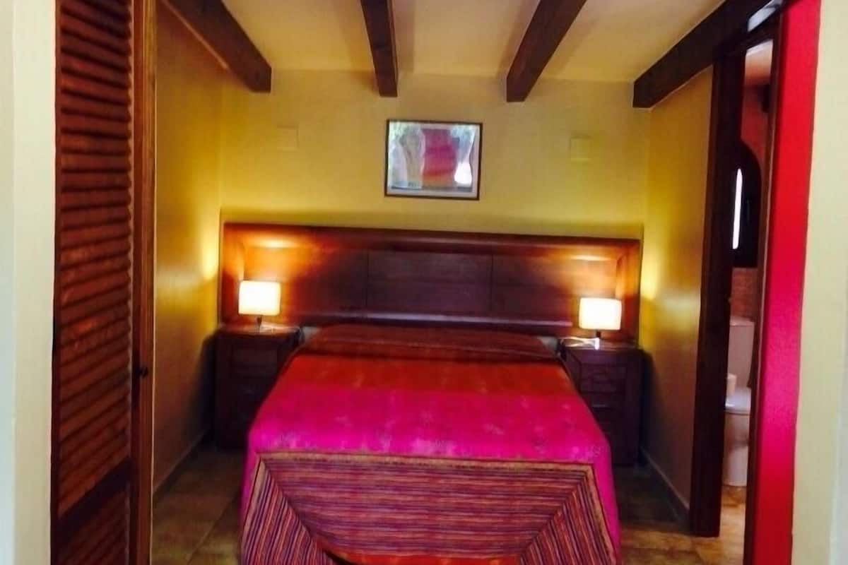 Quarto com cama, luminárias de cabeceira, painel de cabeceira de madeira e uma pequena janela.