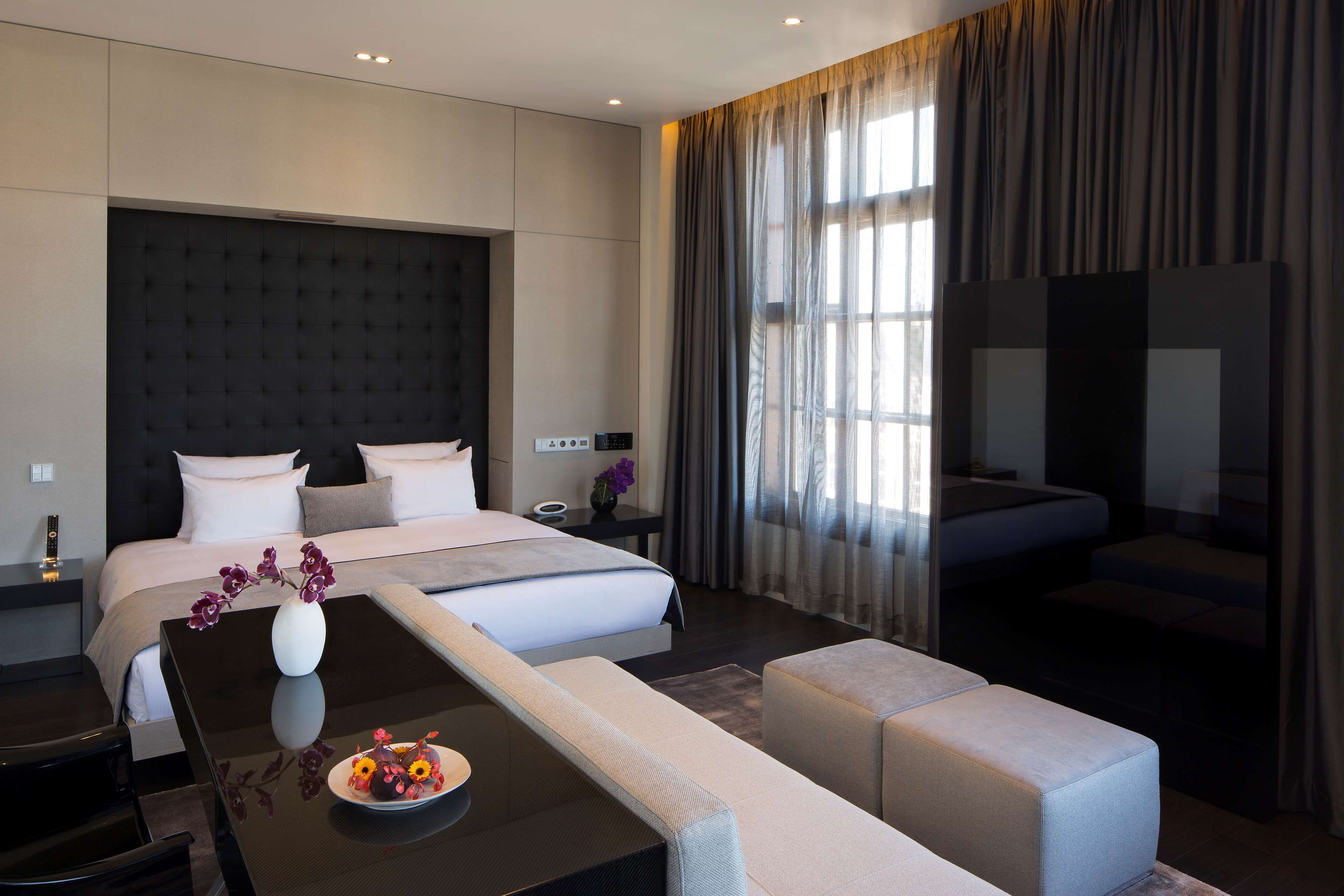 Quarto de hotel moderno com uma cama grande, um sofá, uma mesa de jantar com um vaso de flores e uma televisão.