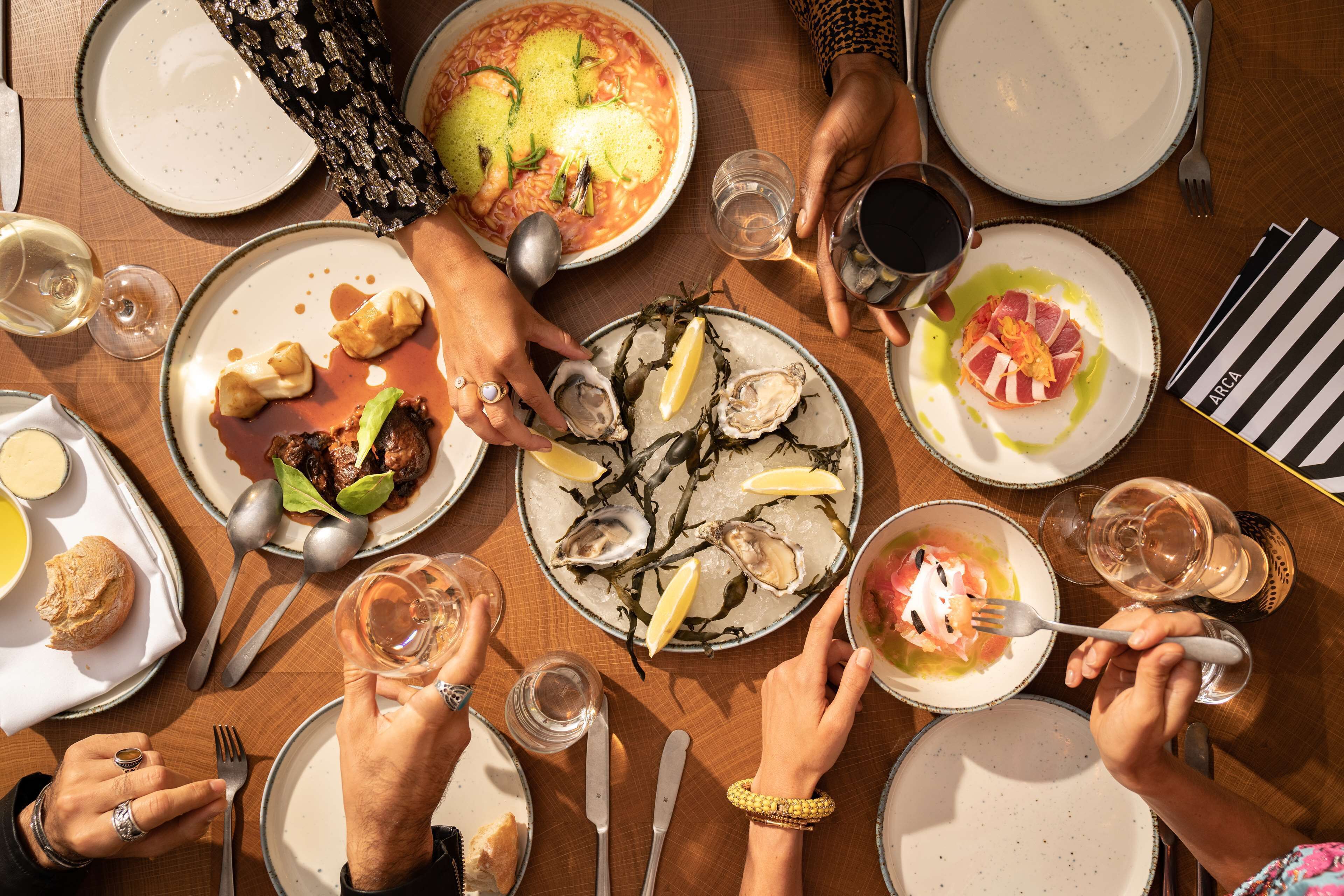 Um grupo de pessoas jantando à mesa, com diversos pratos, incluindo frutos do mar e massa, e várias taças de vinho.