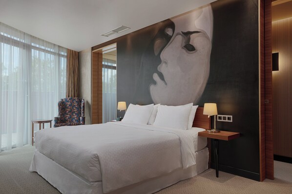 Quarto de hotel moderno com uma cama grande, uma mesa de cabeceira com abajur, uma cadeira e um mural na parede.