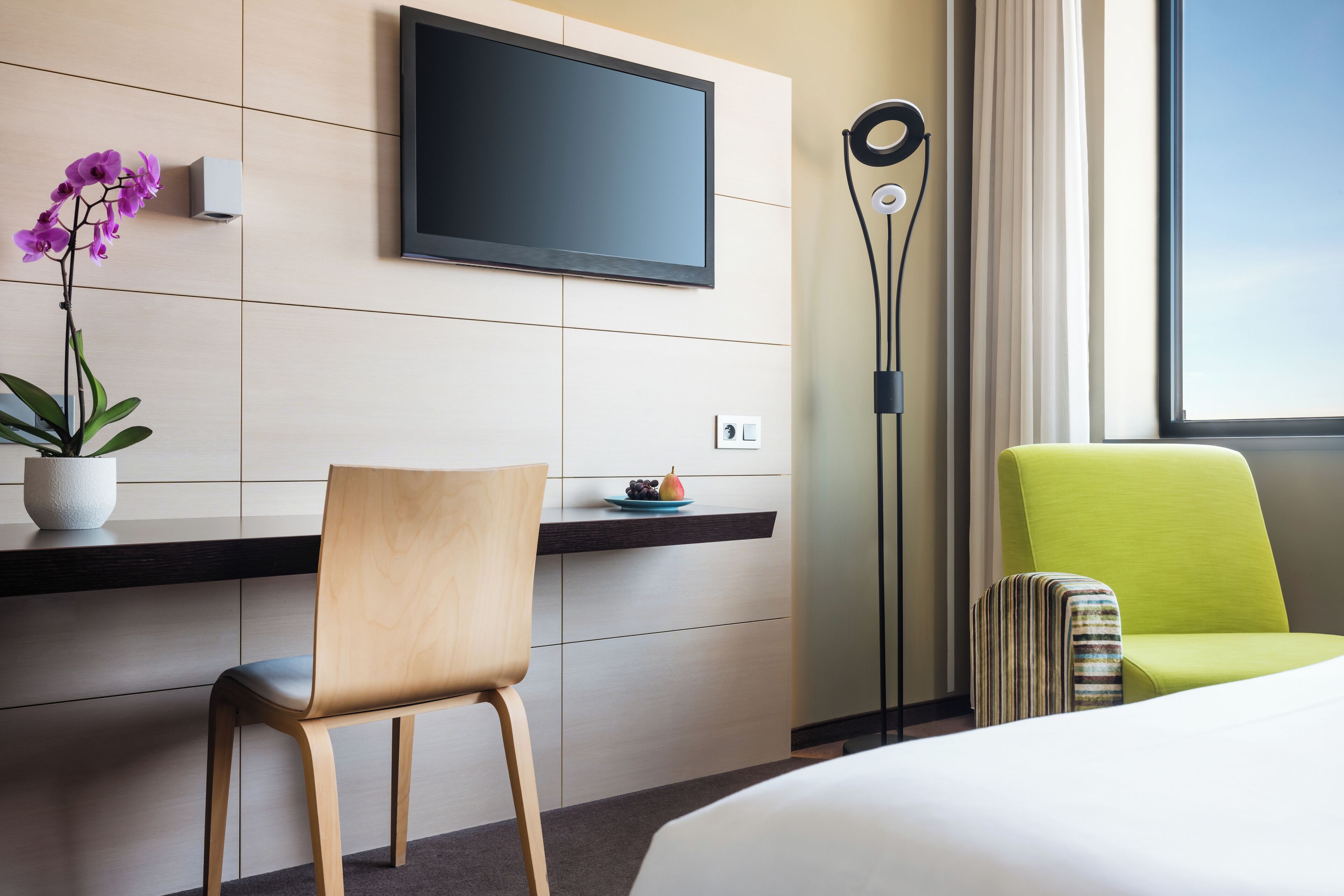 Quarto de hotel com cama, escrivaninha com cadeira, televisão na parede, planta em vaso e janela com cortinas.
