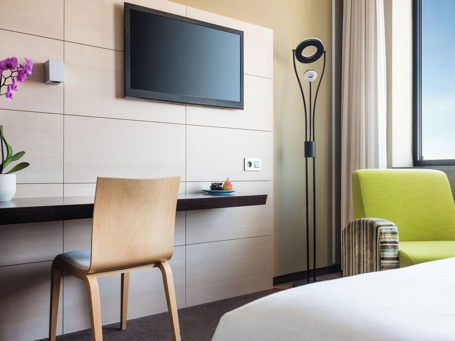 Quarto de hotel com cama, escrivaninha com cadeira, televisão na parede, planta em vaso e janela com cortinas.