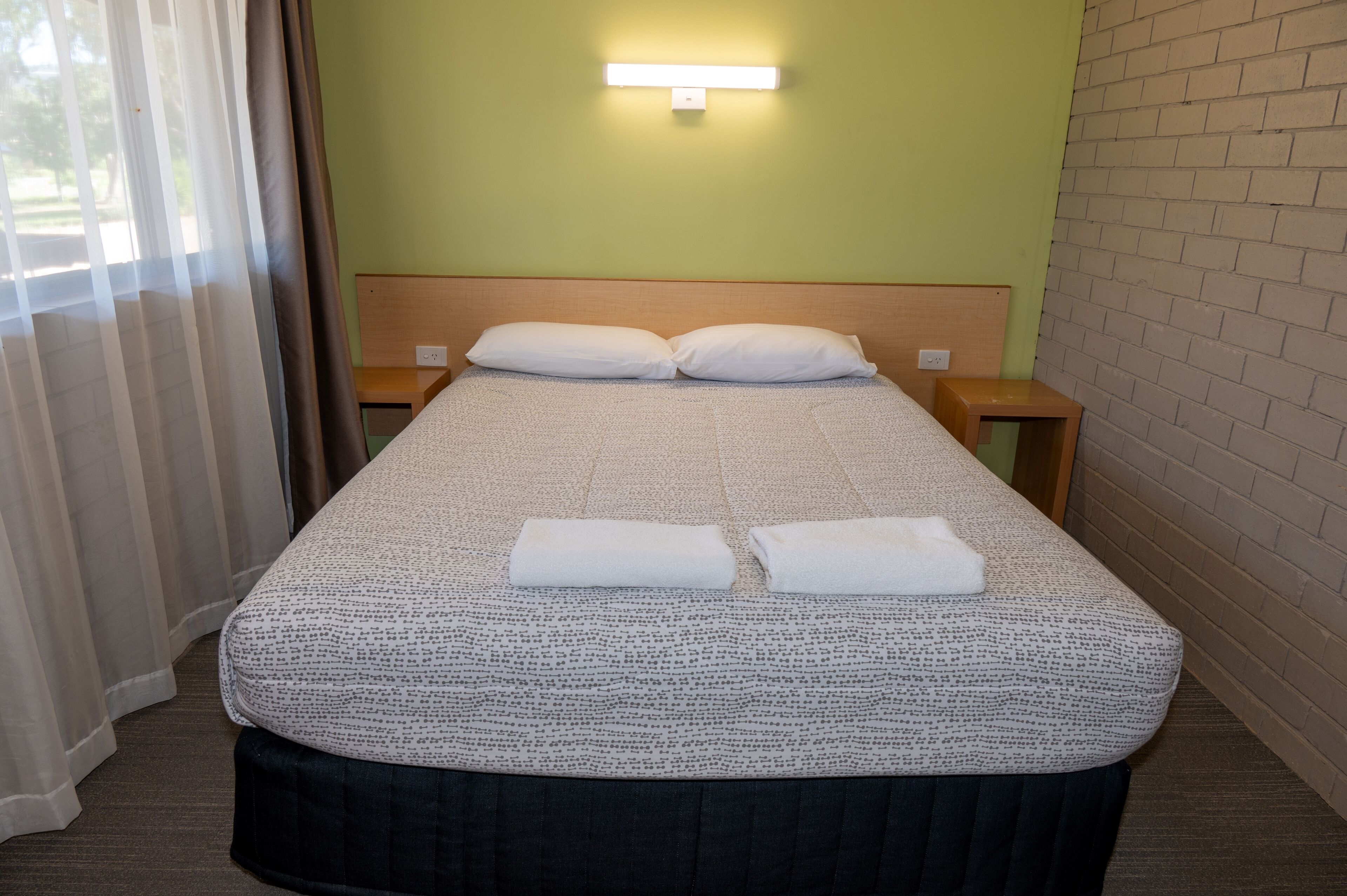 Cama bem arrumada com colcha estampada, duas almofadas e duas toalhas dobradas. Um criado-mudo à direita e uma janela com cortinas à esquerda.