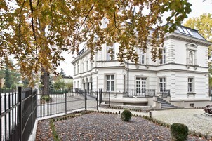 Um edifício grande e branco com fachada simétrica, entrada central com degraus e pequena área de jardim com cascalho e algumas plantas.