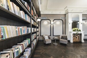 Uma biblioteca com estantes, dois poltronas, uma lareira e uma pequena mesa.