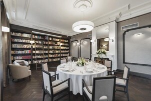 Uma área de refeições estilo biblioteca, com uma mesa redonda posta para seis pessoas, rodeada por estantes repletas de livros.