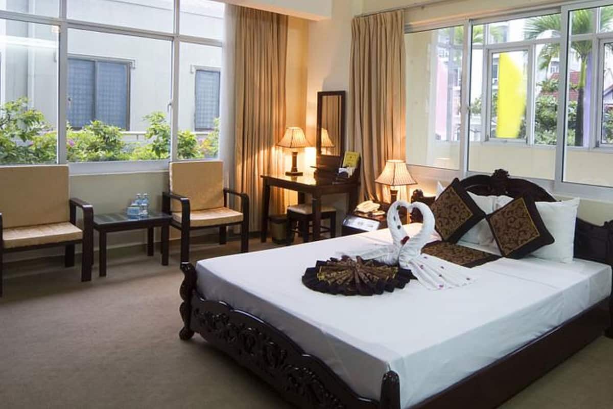 Quarto de hotel com uma cama grande, duas cadeiras, uma escrivaninha e um abajur. Há janelas com cortinas e vista para vegetação lá fora.
