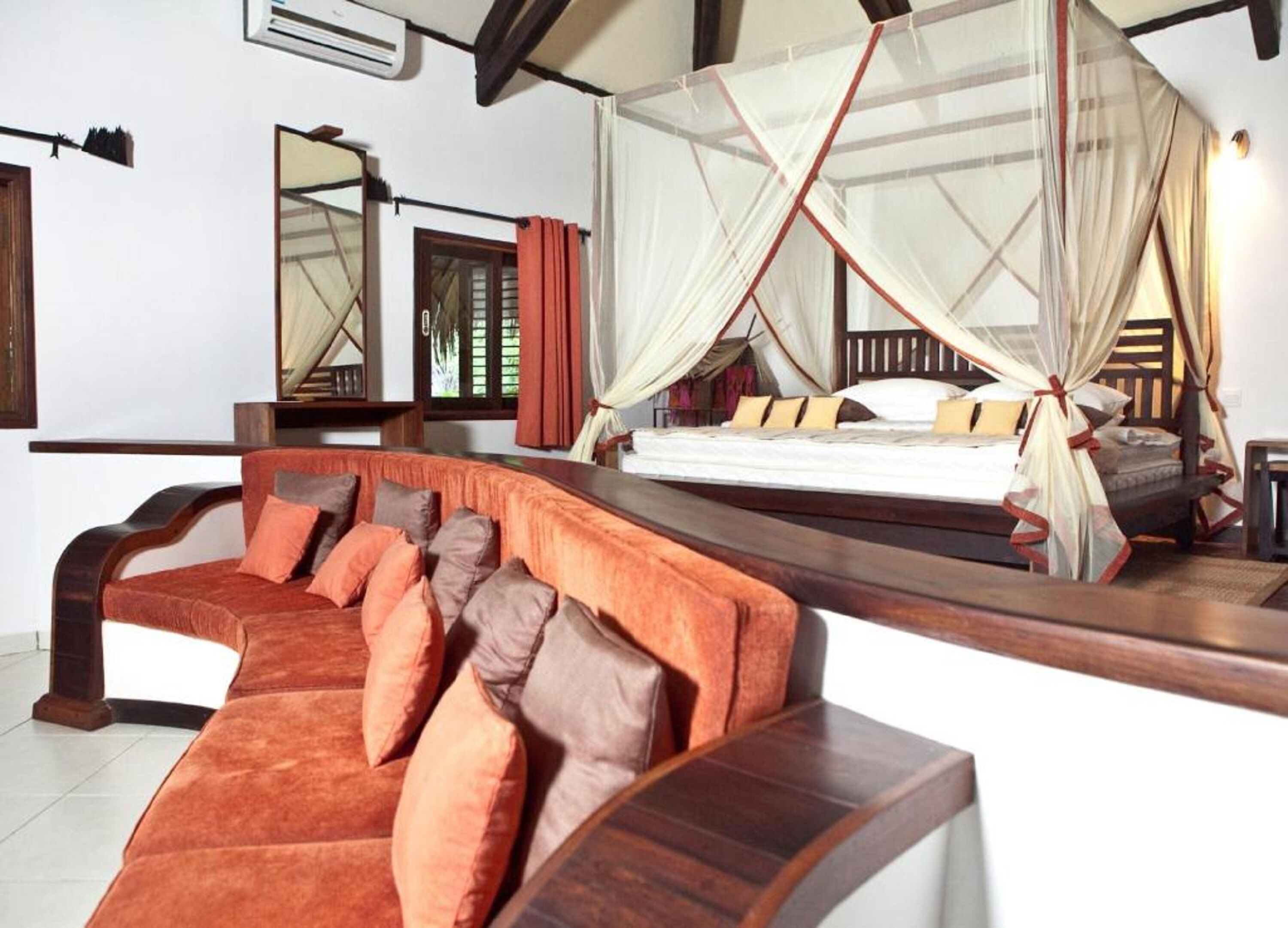 Quarto com uma cama sob um mosquiteiro, um sofá de madeira com almofadas e um espelho grande na parede.