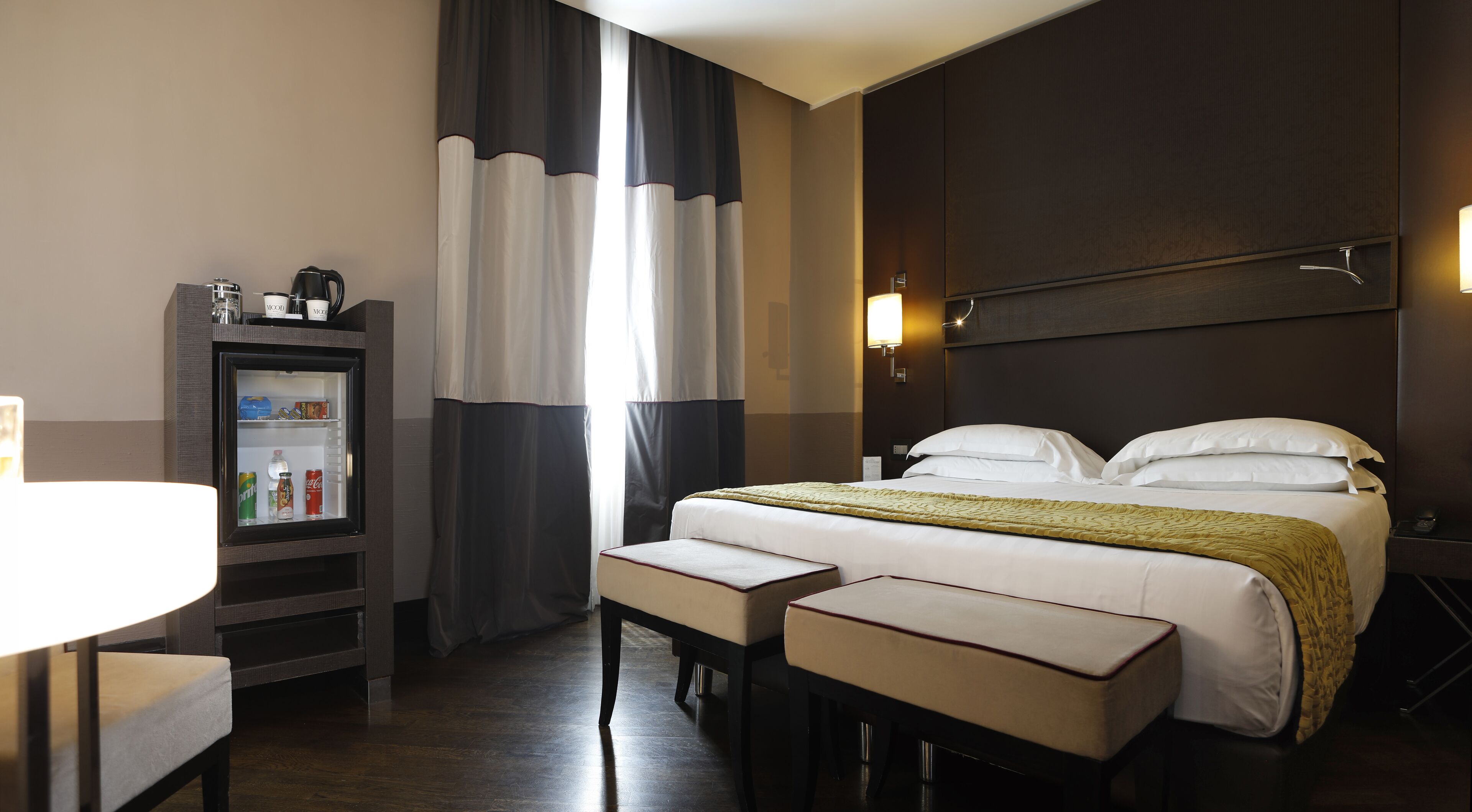 Quarto de hotel com cama, mesas de cabeceira, minibar e escrivaninha com cadeira.