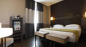 Quarto de hotel com cama, mesas de cabeceira, minibar e escrivaninha com cadeira.