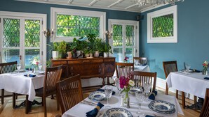 Uma sala de jantar com mesas preparadas para seis pessoas, cada uma com pratos azul e branco, talheres e copos. Há plantas em vasos sobre um armário de madeira, e o espaço possui janelas amplas com vista para o verde lá fora.