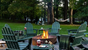 Uma fogueira rodeada por cadeiras Adirondack verdes, uma rede e uma mesa com caneca e marshmallows.
