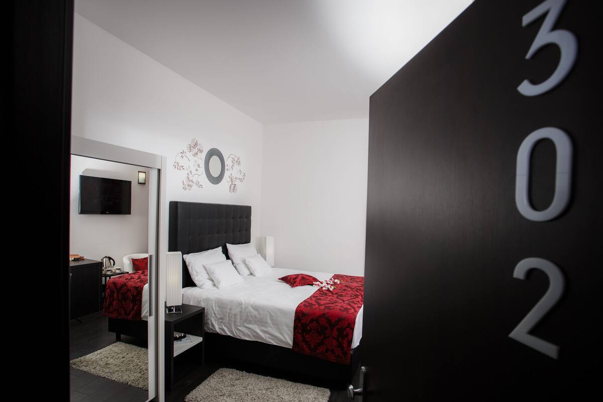 Quarto de hotel moderno com uma cama grande, mesa de cabeceira, televisão na parede e uma porta com números.