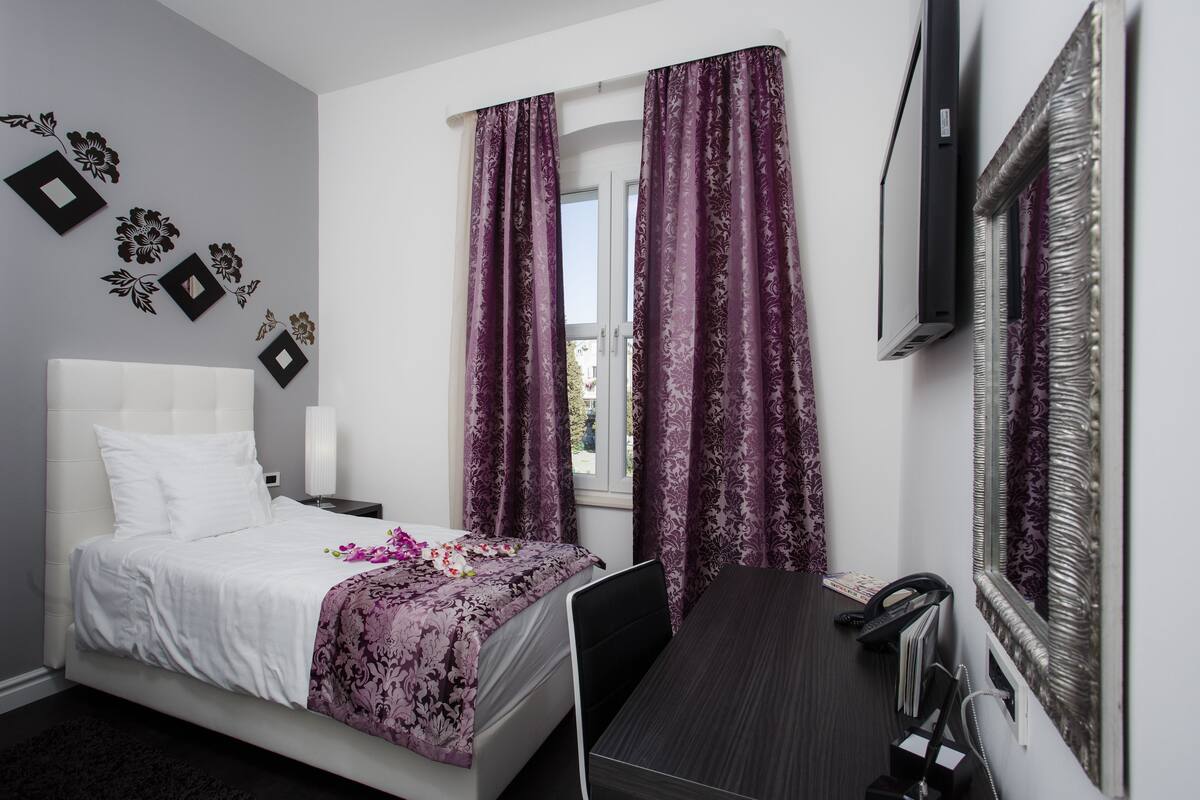 Quarto de hotel moderno com cama, escrivaninha, espelho e janela com cortinas roxas.