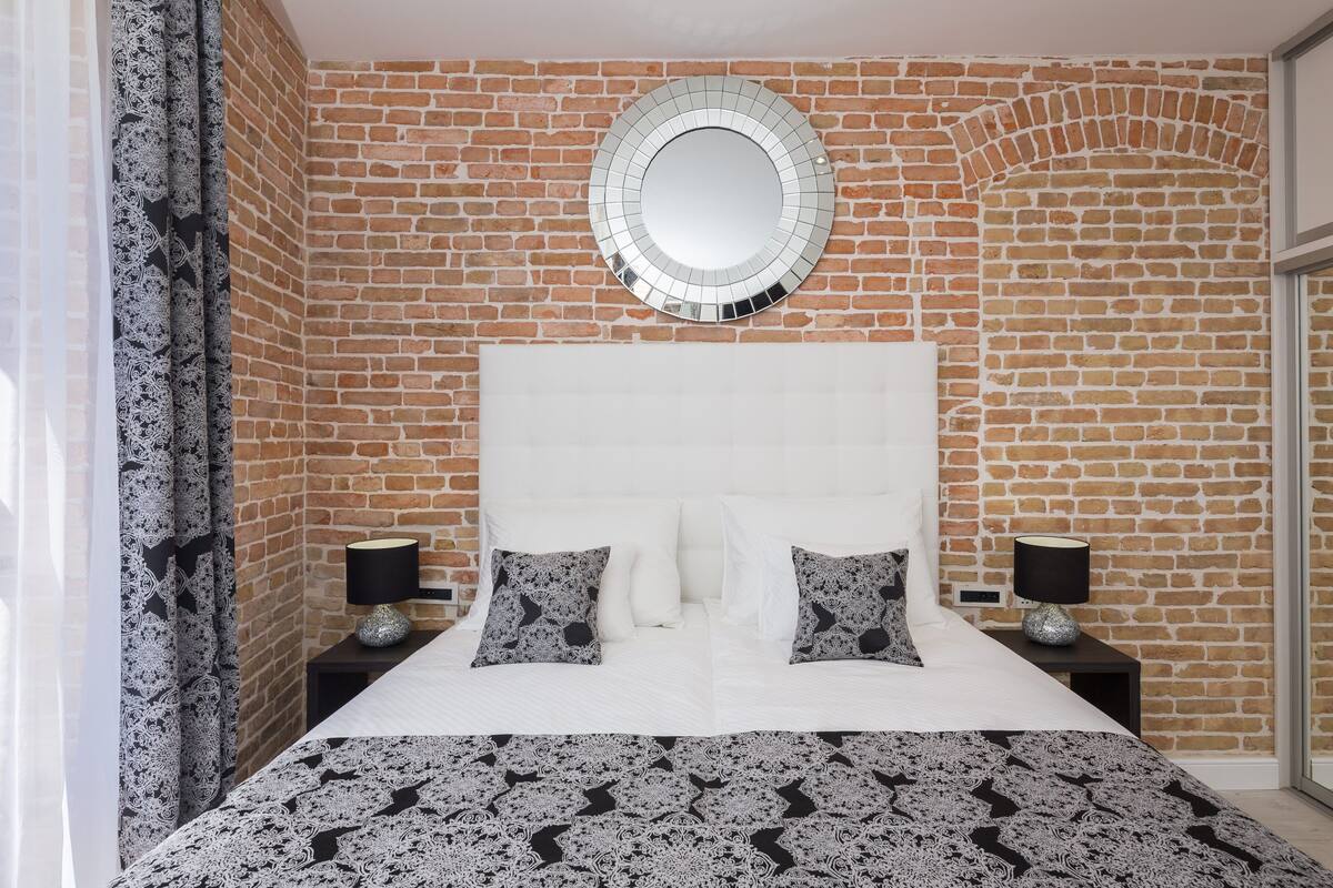 Quarto de hotel com parede de tijolos, espelho redondo, cabeceira branca, colcha estampada, duas mesas de cabeceira com abajures e almofadas estampadas.