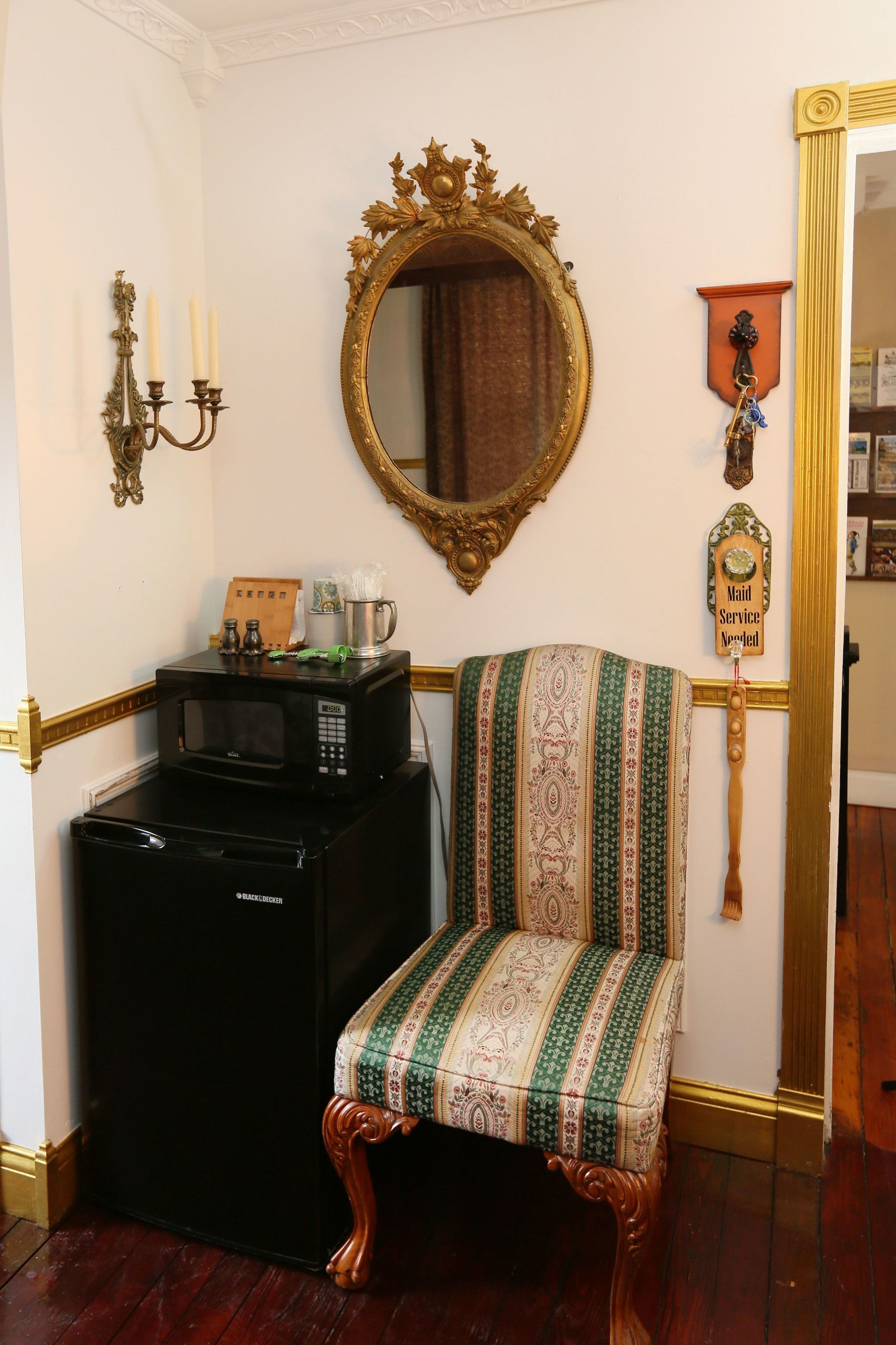 Quarto com geladeira preta, micro-ondas, poltrona estampada, espelho com moldura dourada e cabideiro fixado na parede com uma placa.