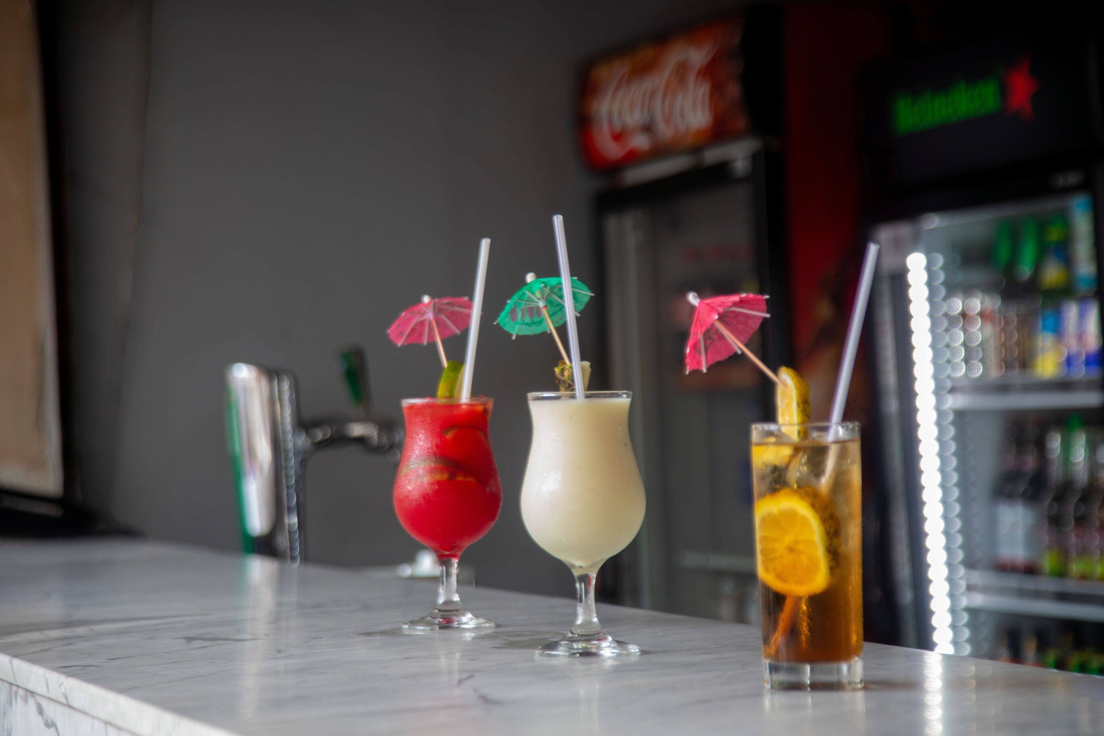 Três bebidas em um balcão de bar, com um fundo desfocado incluindo um letreiro da Coca-Cola.