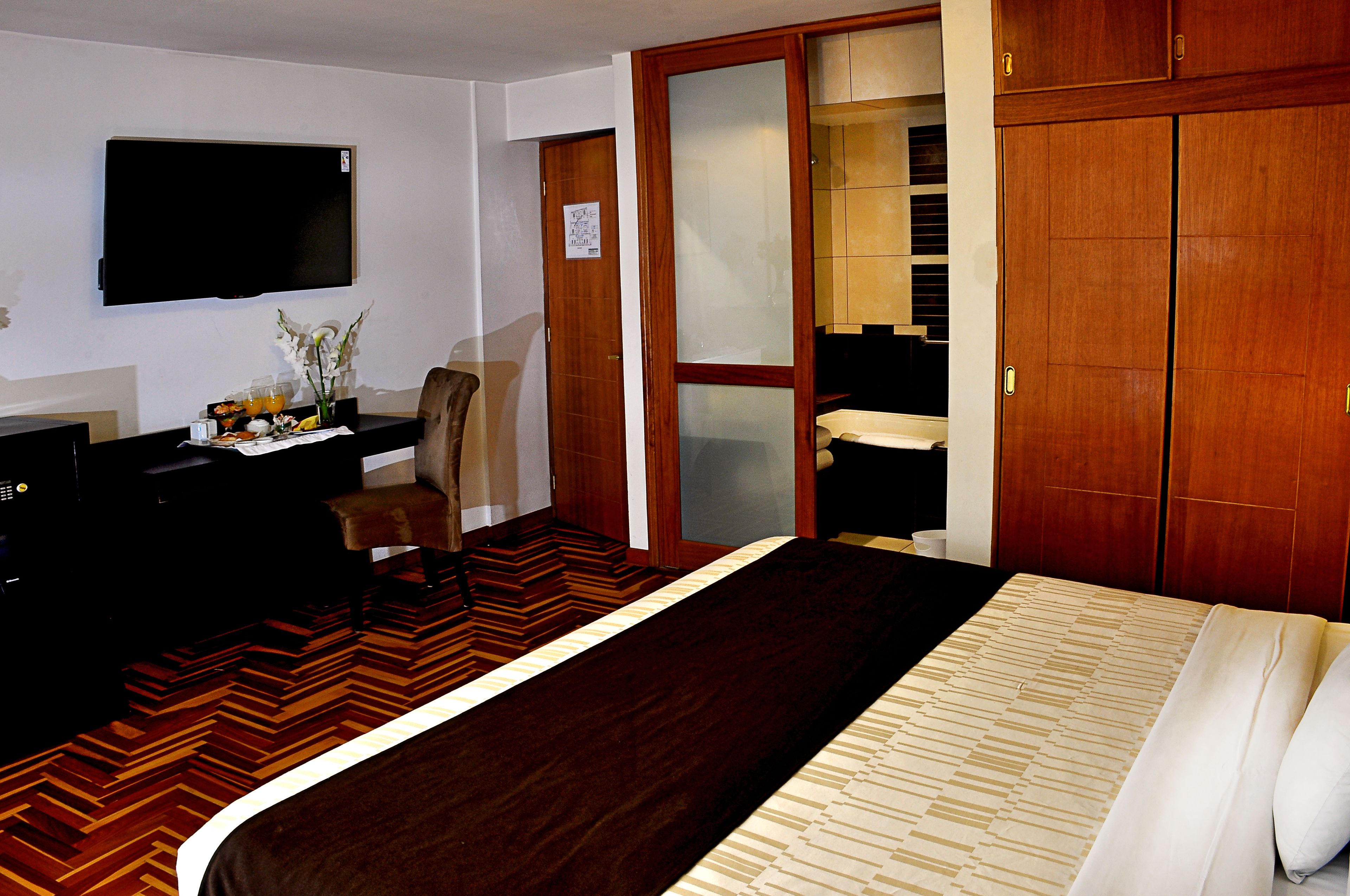 Quarto de hotel com cama, escrivaninha, cadeira, televisão e guarda-roupa.