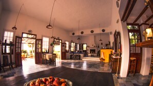 Um quarto espaçoso com uma grande mesa de madeira em primeiro plano, área de cozinha ao fundo, e vista para o exterior através de amplas janelas.
