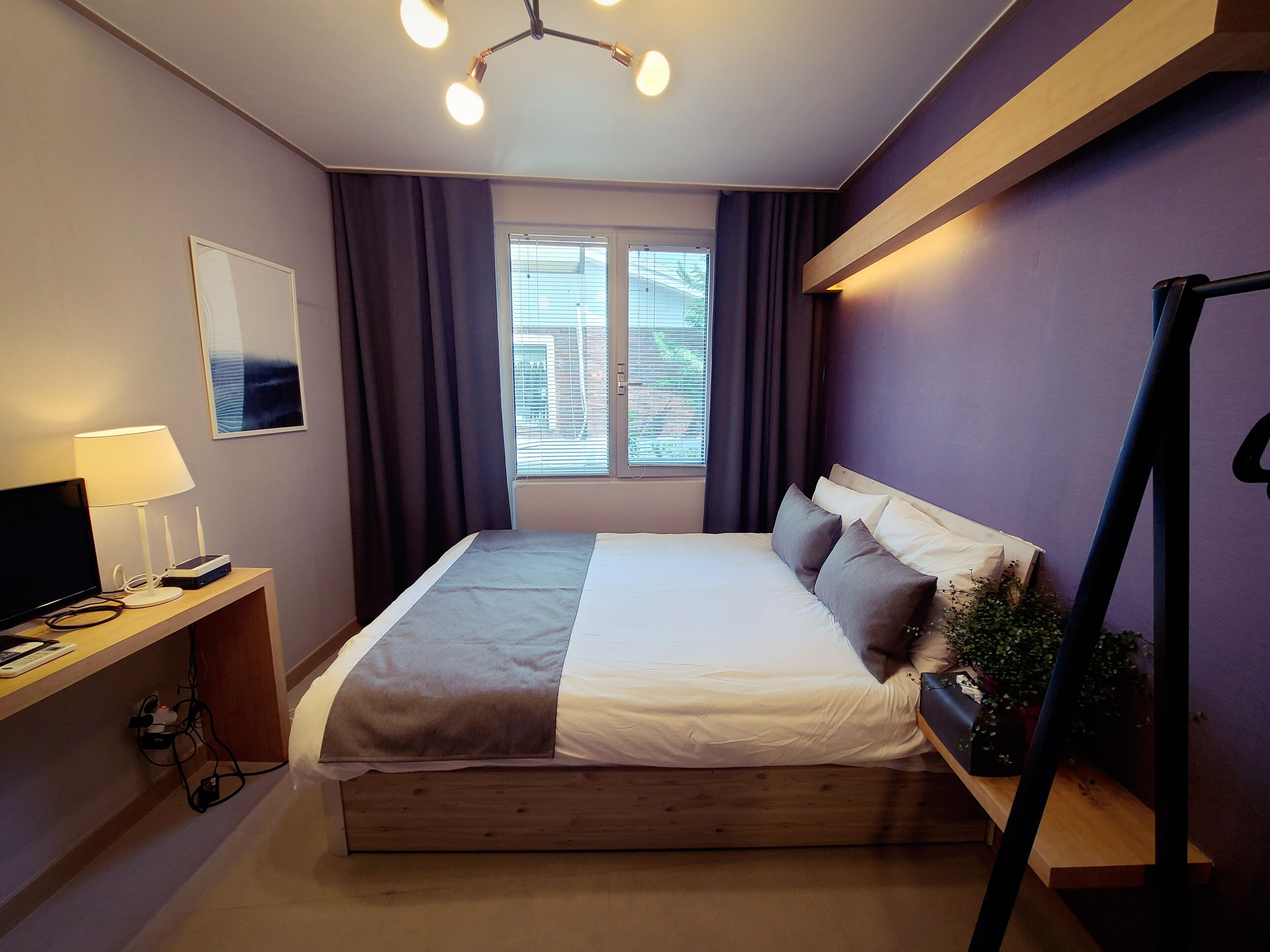 Quarto moderno com cama, escrivaninha com abajur, televisão e janela com cortinas.