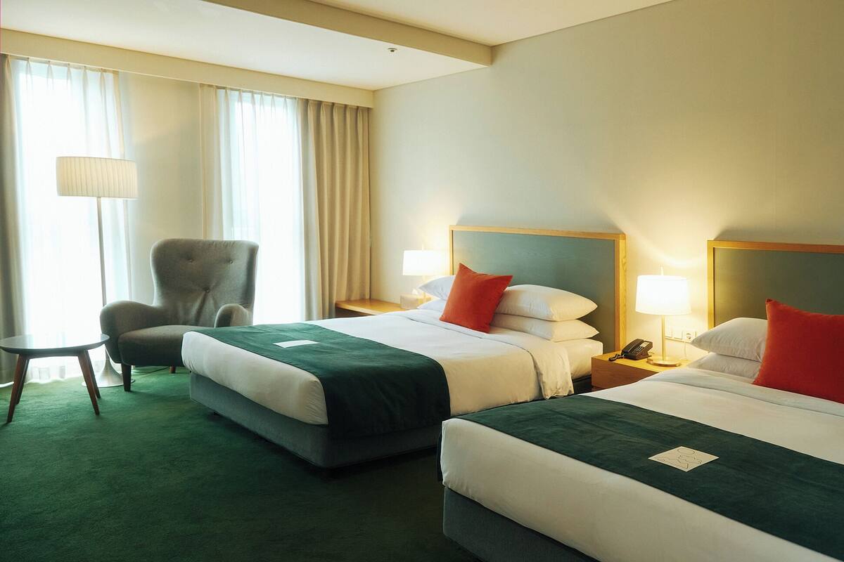 Quarto de hotel com duas camas, uma cadeira, um abajur e uma mesinha de cabeceira.