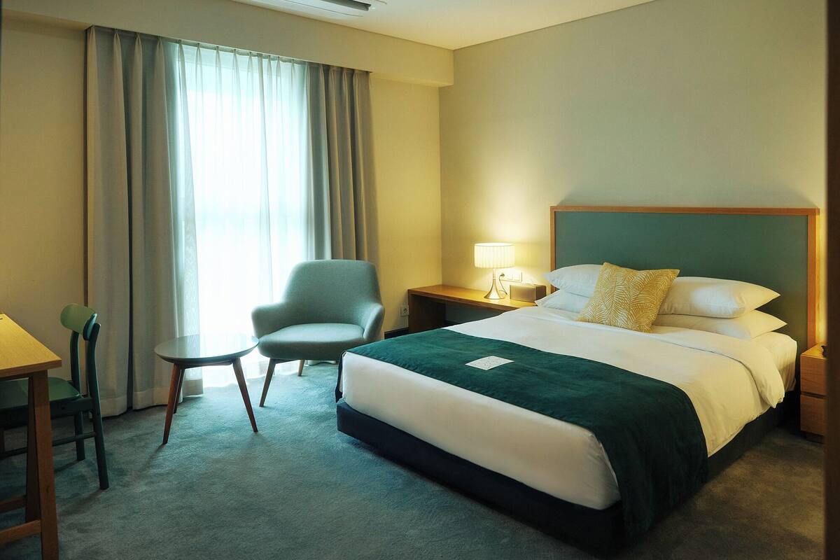 Quarto de hotel com uma cama, uma cadeira, uma mesinha, uma mesa de cabeceira com abajur e uma janela com cortinas.