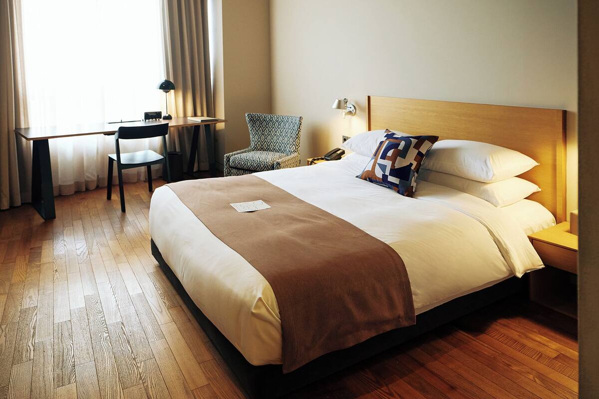 Quarto de hotel com uma cama, uma escrivaninha, uma cadeira e uma mesinha de cabeceira.