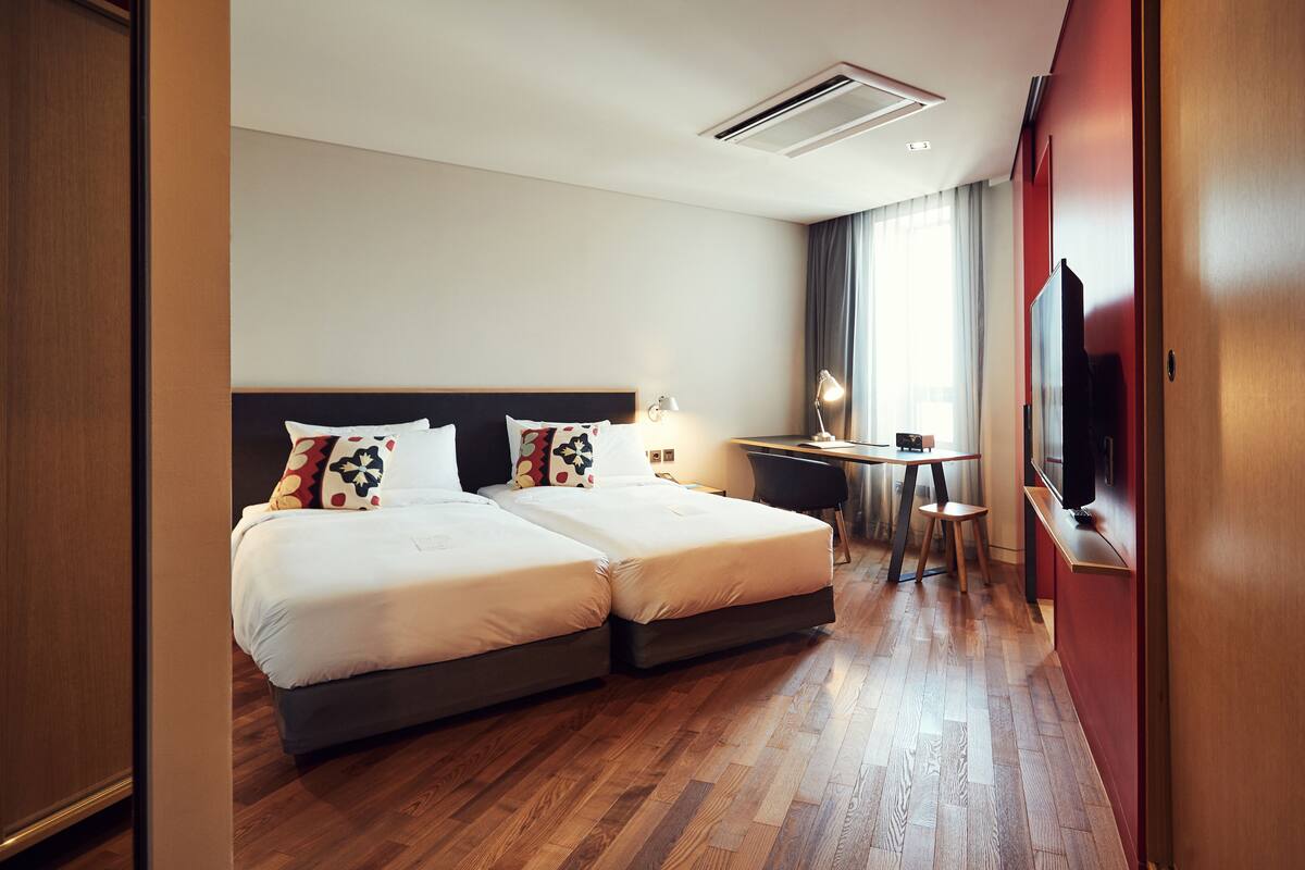 Quarto de hotel com duas camas, uma escrivaninha, uma cadeira, uma televisão e uma janela com cortinas.