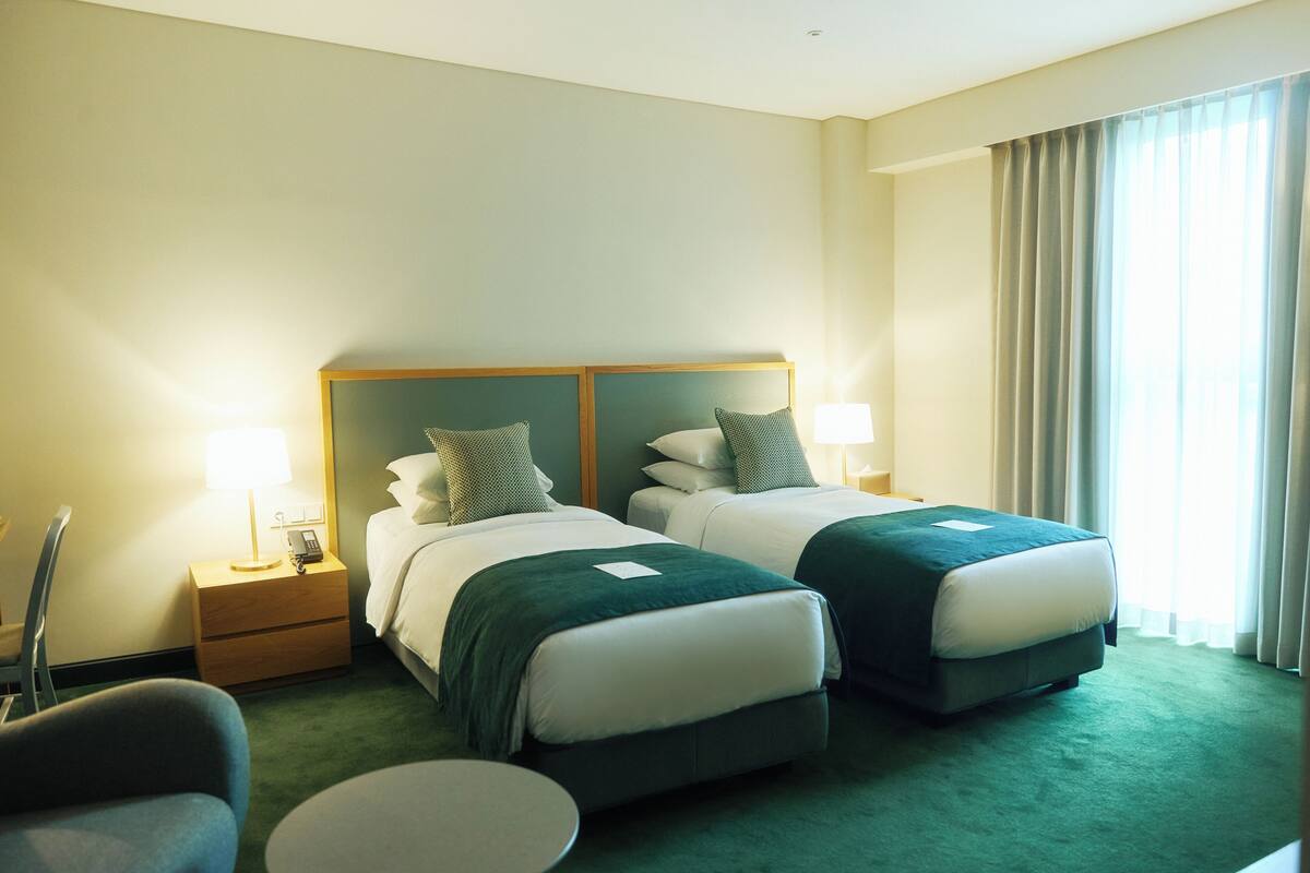 Quarto de hotel com duas camas, uma escrivaninha, uma cadeira e uma mesa redonda.