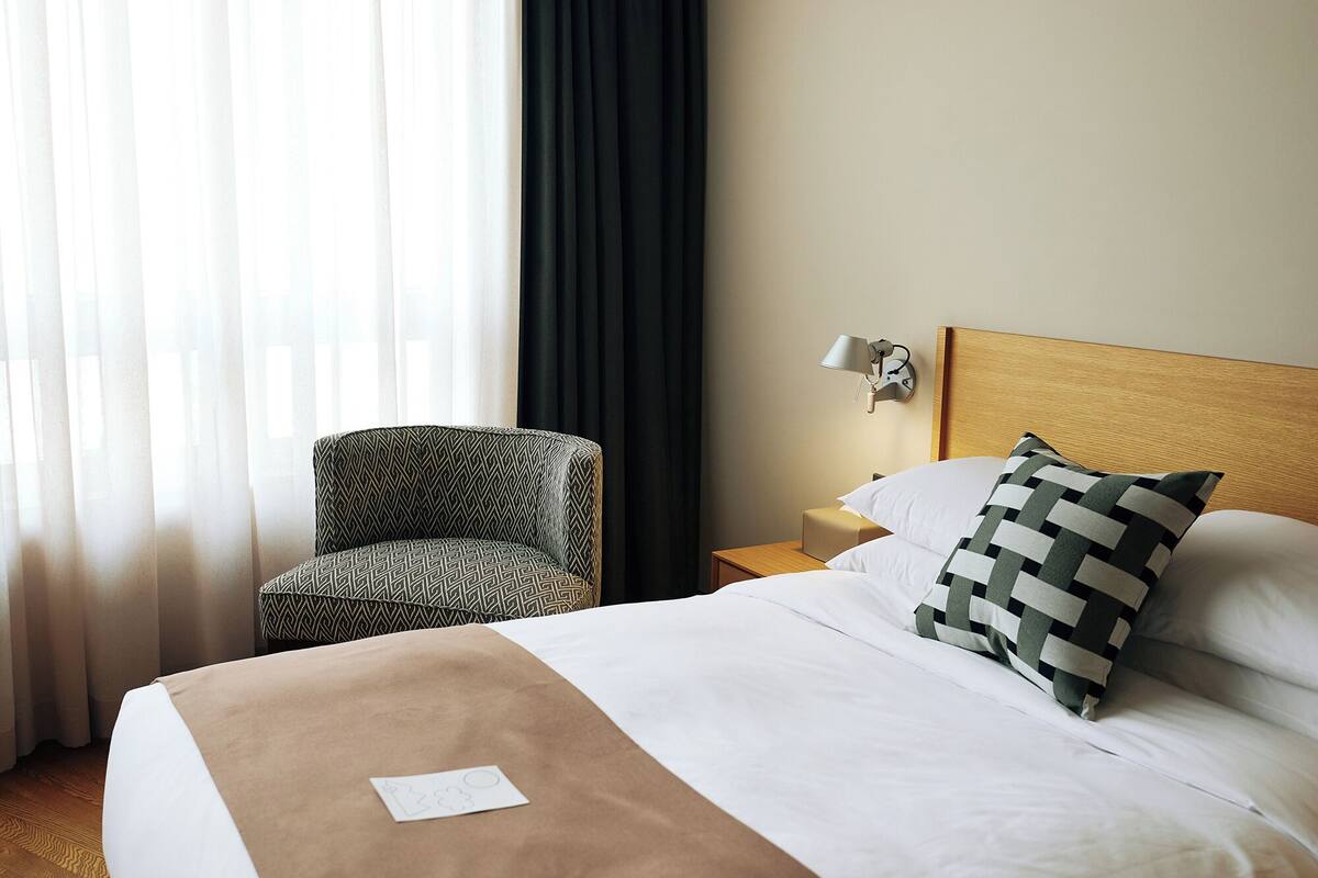 Quarto de hotel com cama, poltrona, mesa de cabeceira e luminária.