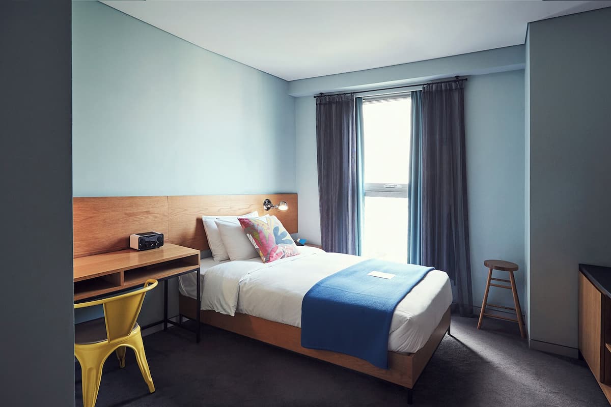 Quarto de hotel com uma cama, uma escrivaninha, uma cadeira, uma televisão e uma janela com cortinas.